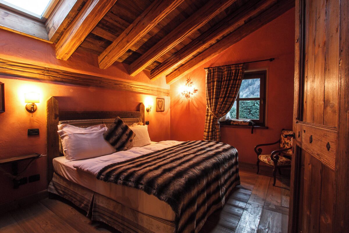 Quarto aconchegante com teto de madeira, uma cama com manta listrada, mesa de cabeceira, uma cadeira e uma janela com cortinas.