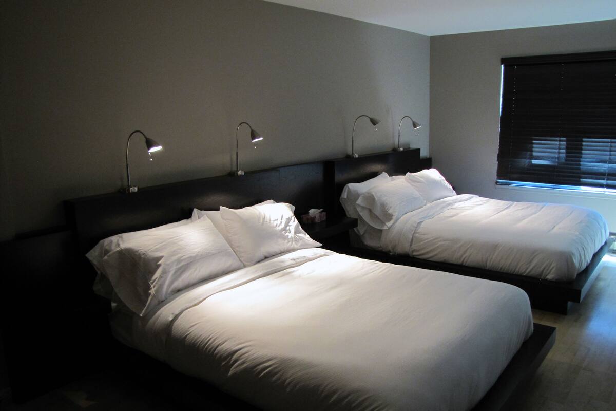 Quarto de hotel com duas camas, cada uma com lençóis brancos e cabeceiras escuras. Três abajures na parede.