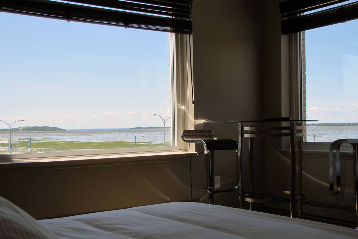 Quarto de hotel com cama, poltrona, mesa e janela com vista para um corpo de água e uma linha de costa distante.