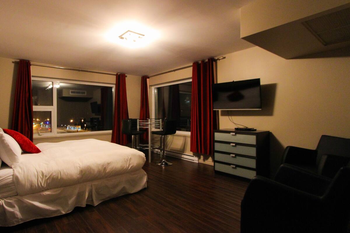 Quarto de hotel com cama, escrivaninha, cadeira, televisão e janela com cortinas.