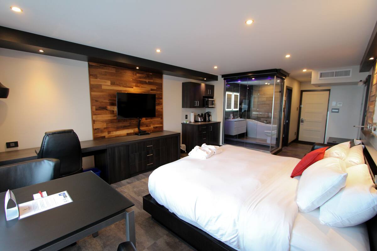 Quarto de hotel moderno com uma cama grande, uma mesa com TV de tela plana, um painel de parede em madeira e banheiro separado com chuveiro de vidro.