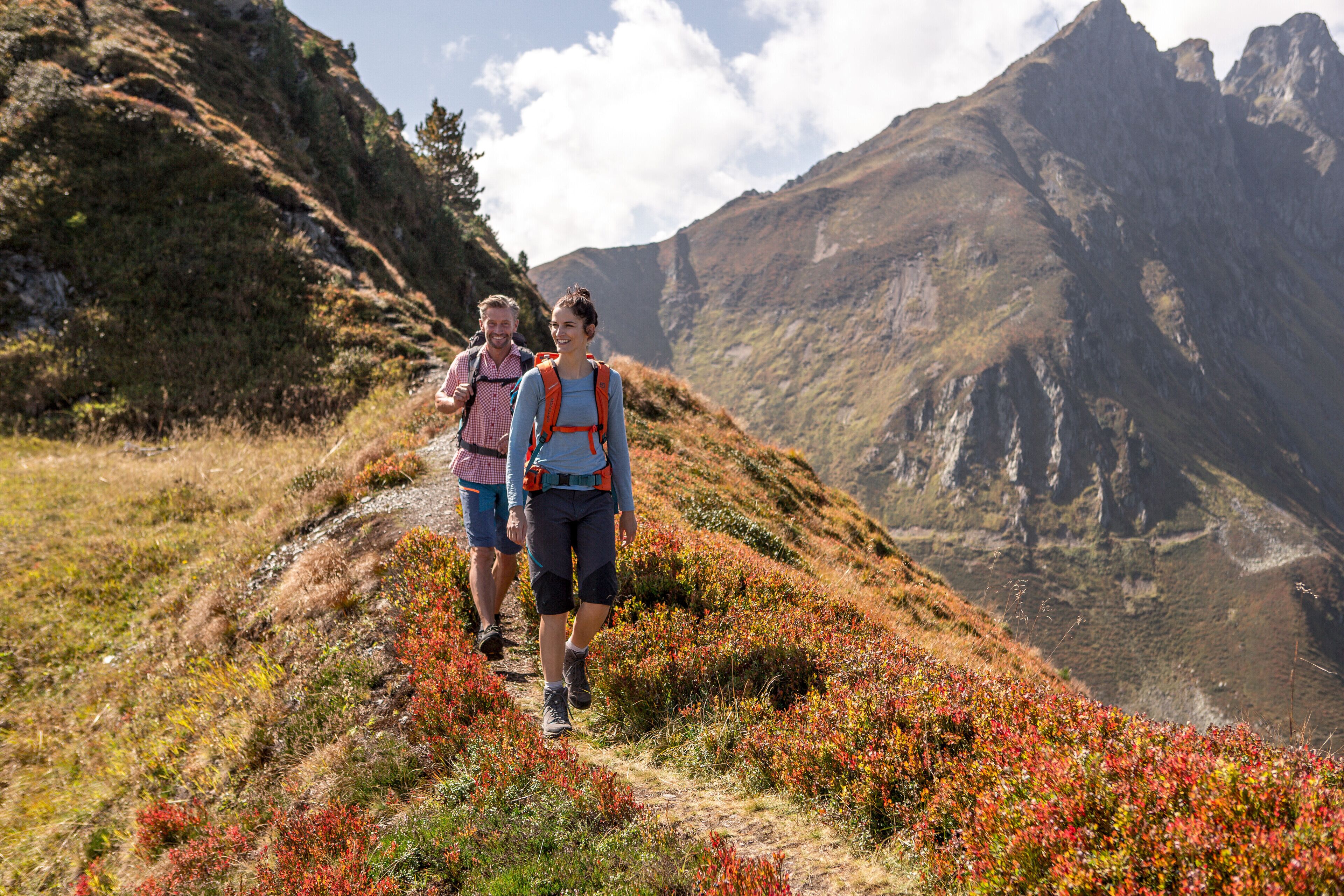Duas pessoas caminhando por uma trilha montanhosa com terreno rochoso e vegetação escassa.