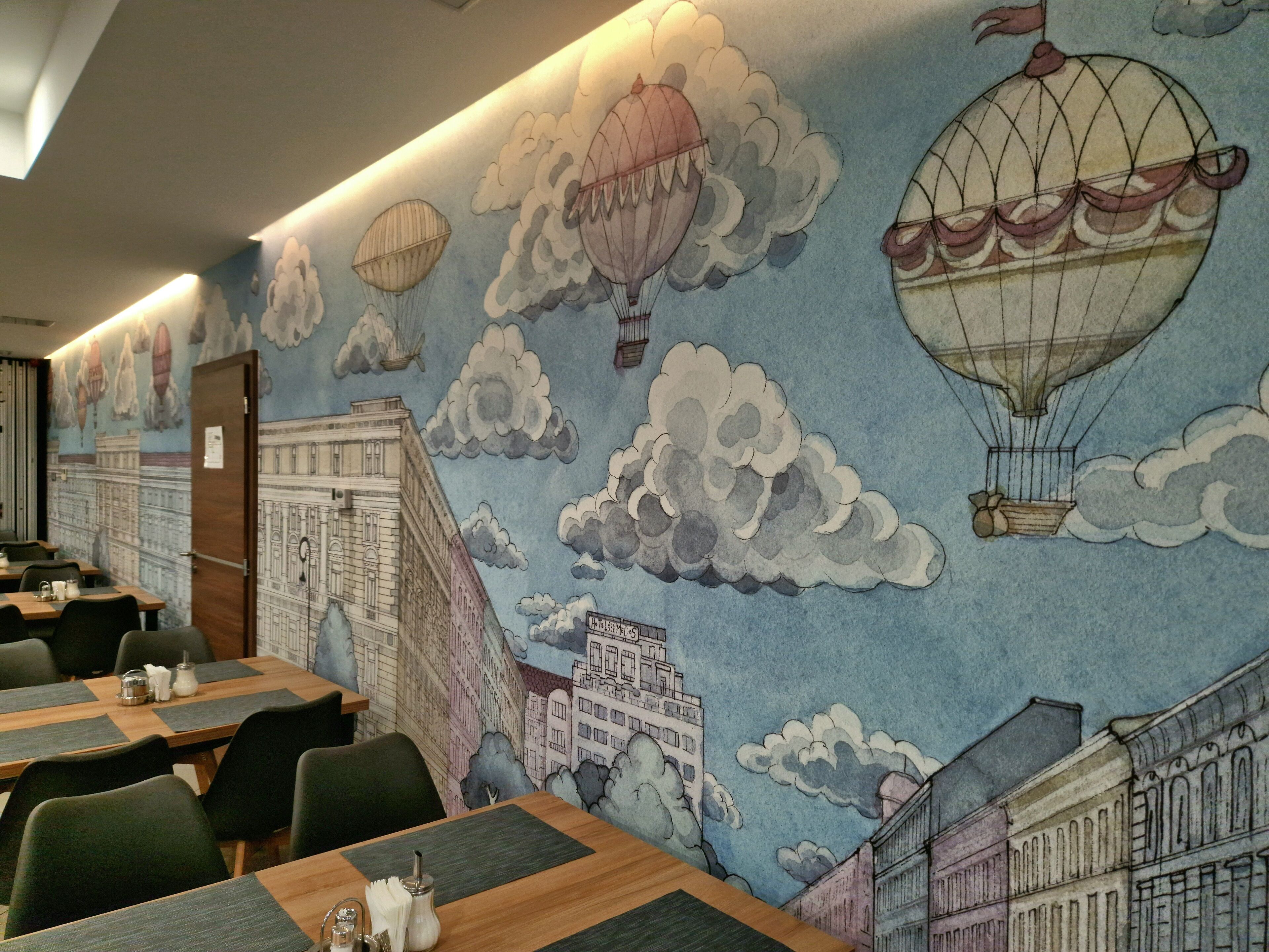 Restaurante com um mural de balões de ar quente e edifícios. Mesas dispostas com cadeiras.