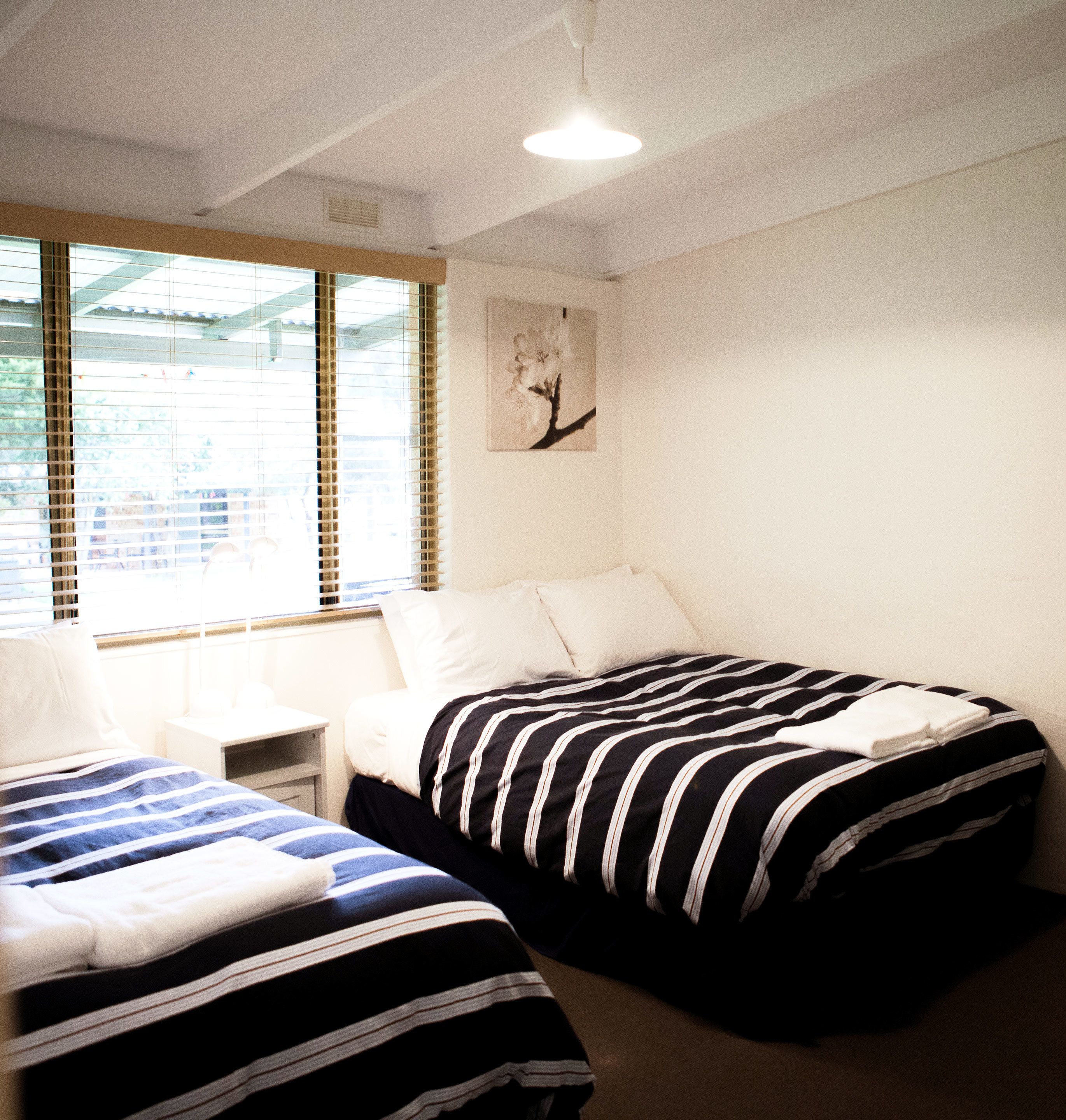 Quarto de hotel com duas camas, uma mesinha de cabeceira, janela com persianas, luminária no teto e um quadro pendurado na parede.