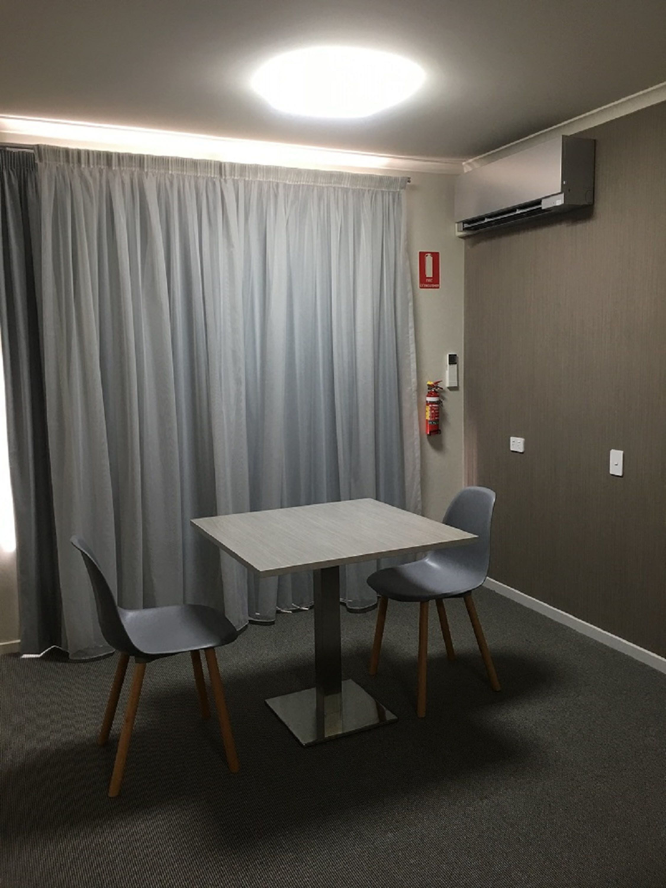 Um quarto pequeno com uma mesa e duas cadeiras, um extintor de incêndio e uma luminária no teto.
