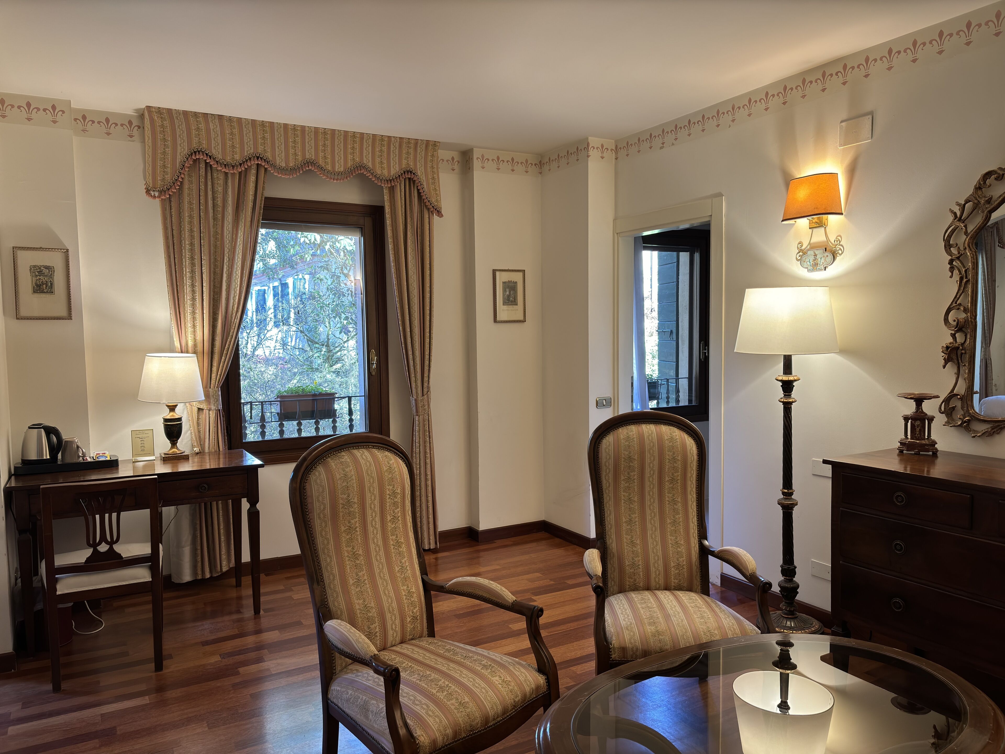Quarto de hotel com escrivaninha de madeira, duas poltronas estofadas, mesa redonda de vidro, penteadeira e um espelho grande.