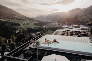 Uma piscina no terraço com duas pessoas em bóias, rodeada por espreguiçadeiras e guarda-sóis, com vista para um vale e montanhas ao fundo.