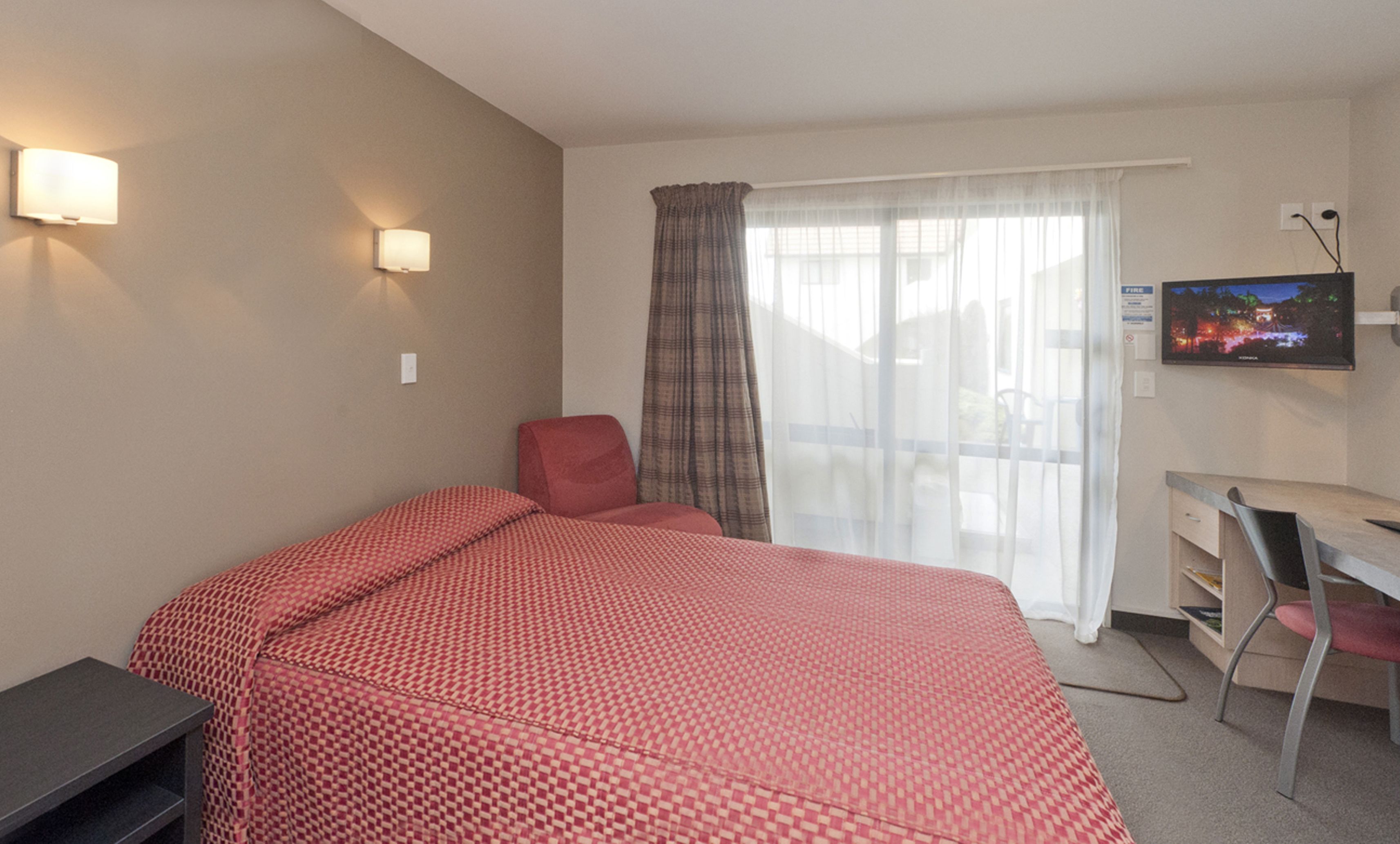 Quarto de hotel com cama, escrivaninha, cadeira, televisão e janela com cortinas.