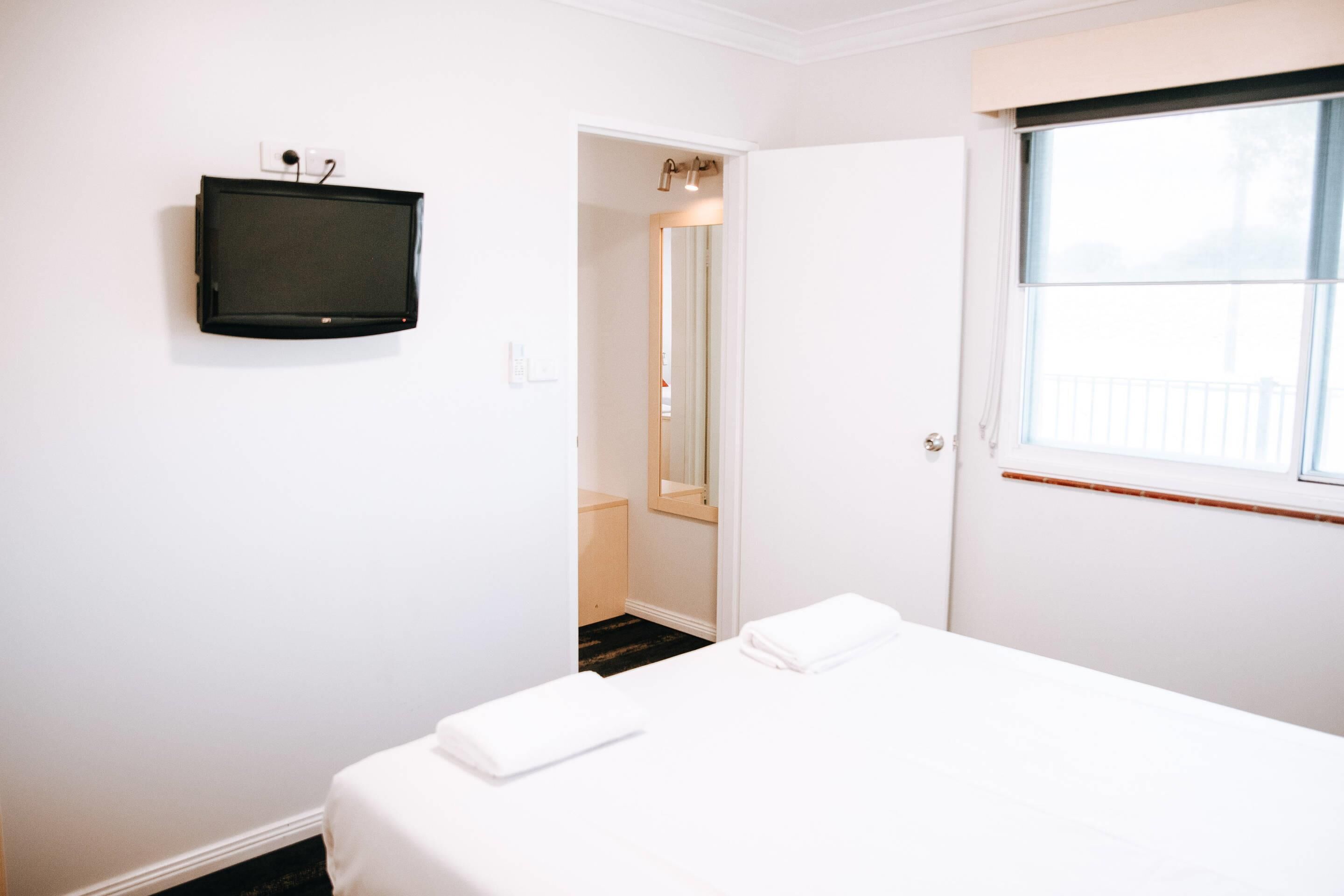 Quarto de hotel com cama, televisão fixada na parede, janela e porta de acesso a outro espaço.