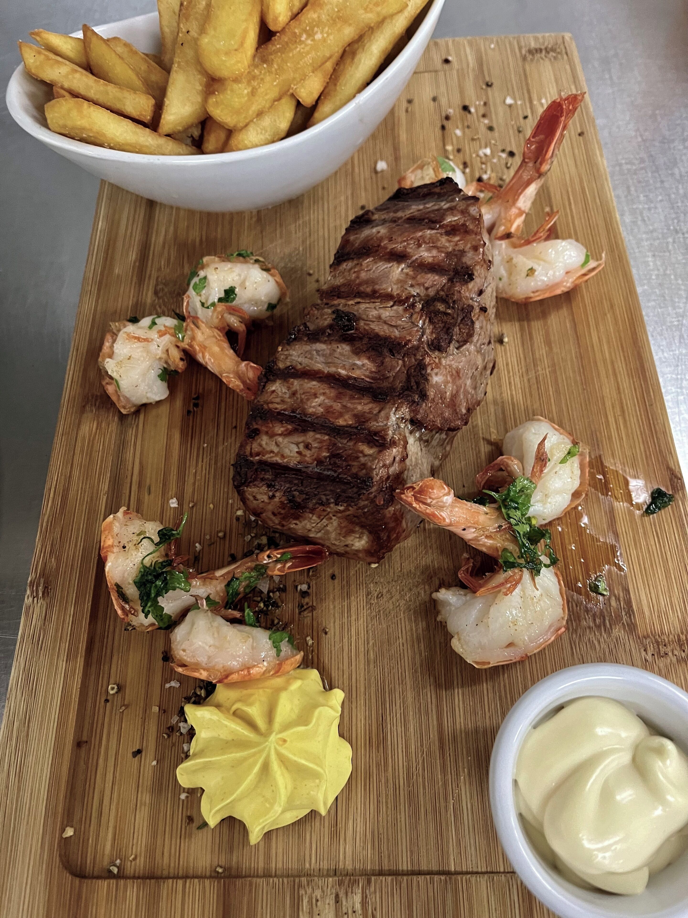 Tábua de madeira com bife grelhado, camarões e uma pequena tigela de molho.