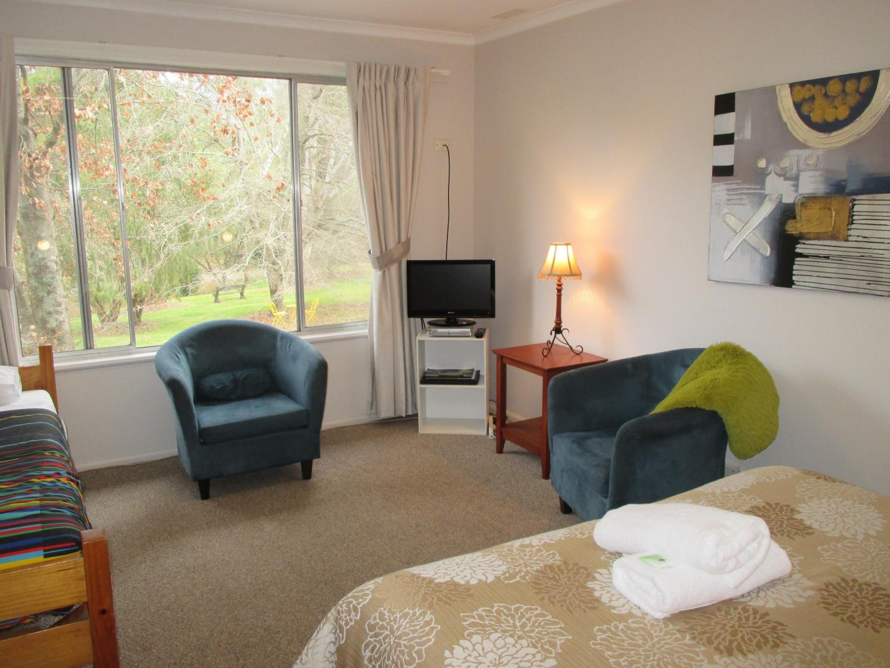 Quarto de hotel com duas poltronas azuis, uma cama com colcha florida, uma televisão sobre um suporte e um abajur sobre uma mesinha.