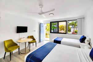 Quarto de hotel com cama, duas mesas de cabeceira, televisão, ventoinha no teto, janela com vista para o verde e uma pequena mesa com uma cadeira.