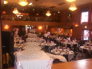 Salão de banquetes com várias mesas redondas preparadas para um evento formal, com uma apresentação central de copos de vinho e balões.