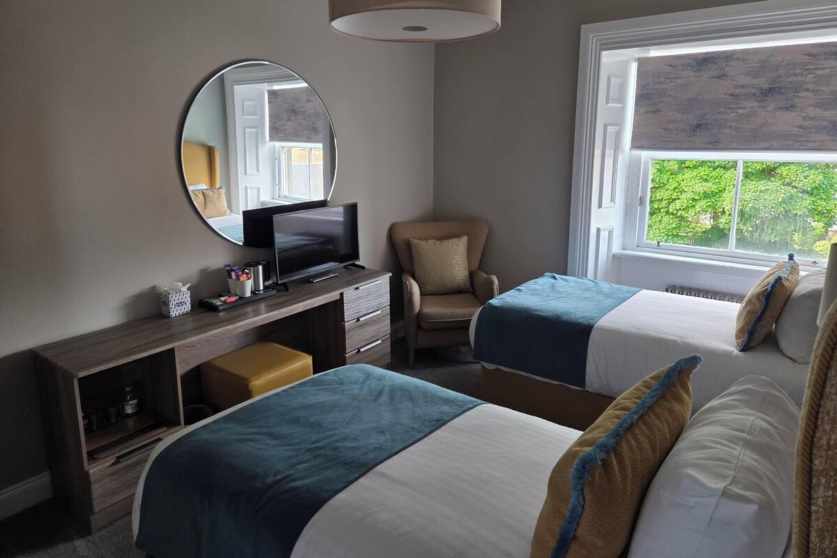 Quarto de hotel com duas camas, uma escrivaninha com televisão, uma cadeira e uma janela ampla com vista para o verde.