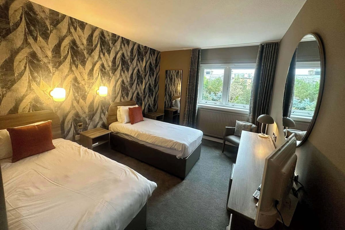 Quarto de hotel com duas camas, uma escrivaninha, uma cadeira, um espelho e uma janela com cortinas.