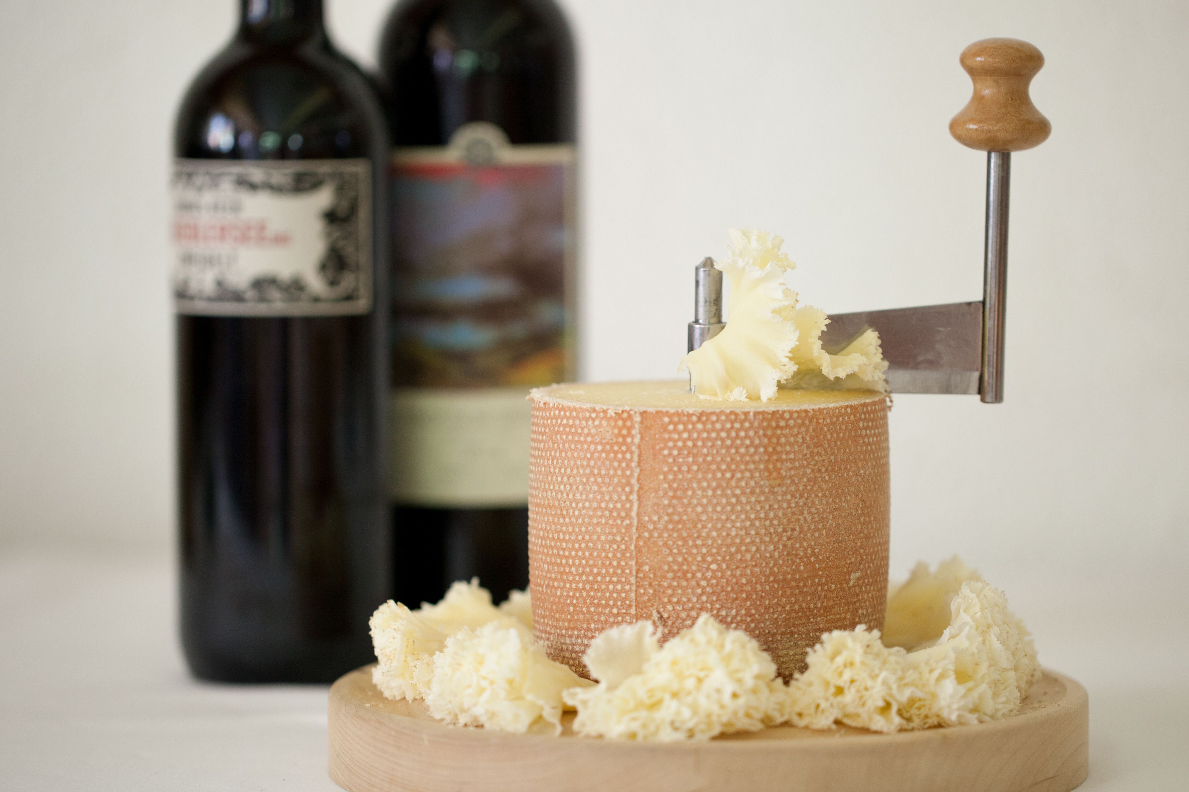 Um ralador de queijo com lascas de queijo sobre uma tábua de madeira, com duas garrafas ao fundo.