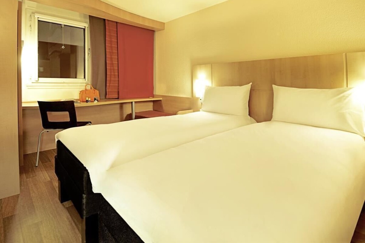 Quarto de hotel com duas camas, uma escrivaninha, uma cadeira e uma janela com cortinas.