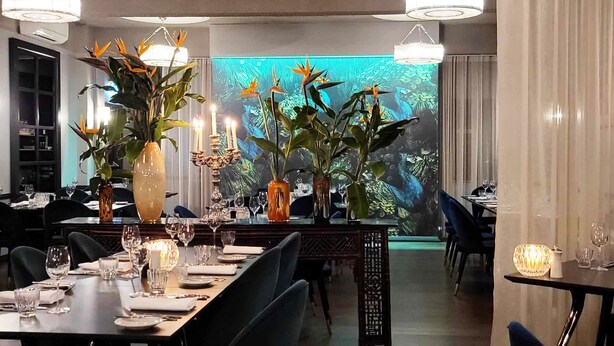 Restaurante com mesas postas para refeições, apresentando grandes vasos com plantas e velas, e uma tela grande exibindo uma cena natural.