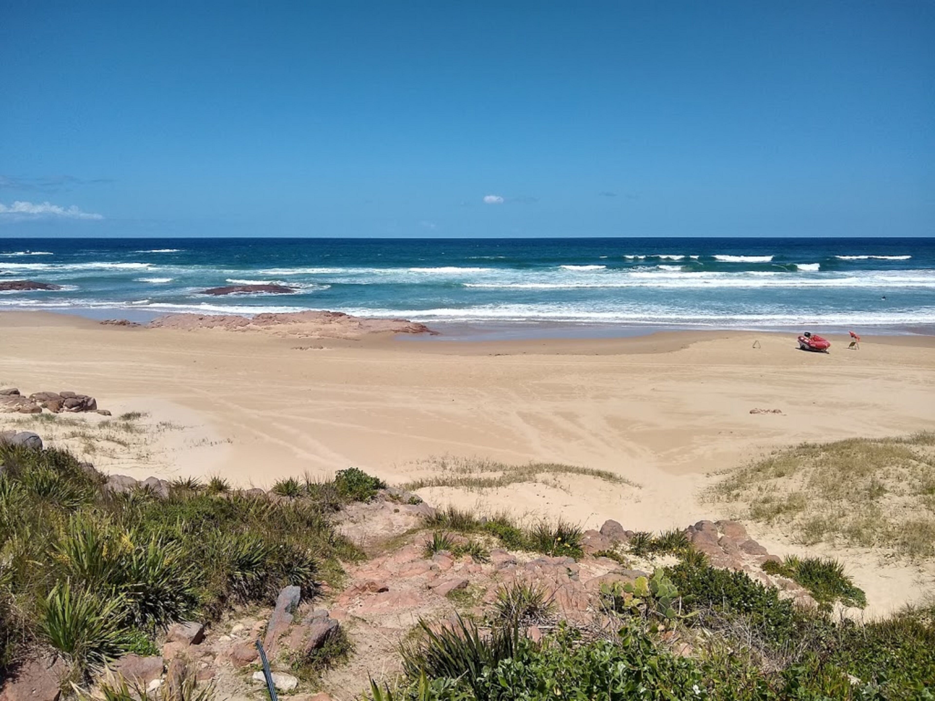 Uma praia com céu claro, areal arenoso e afloramentos rochosos.
