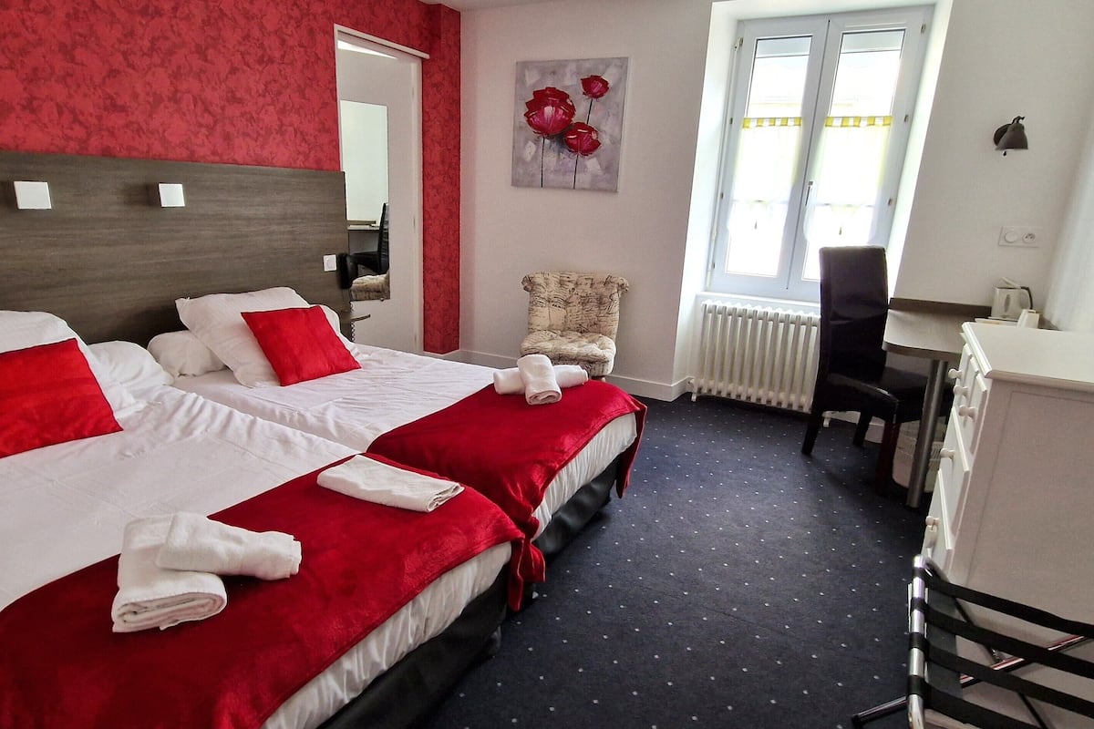 Quarto de hotel com duas camas, uma cadeira, uma escrivaninha e um radiador.