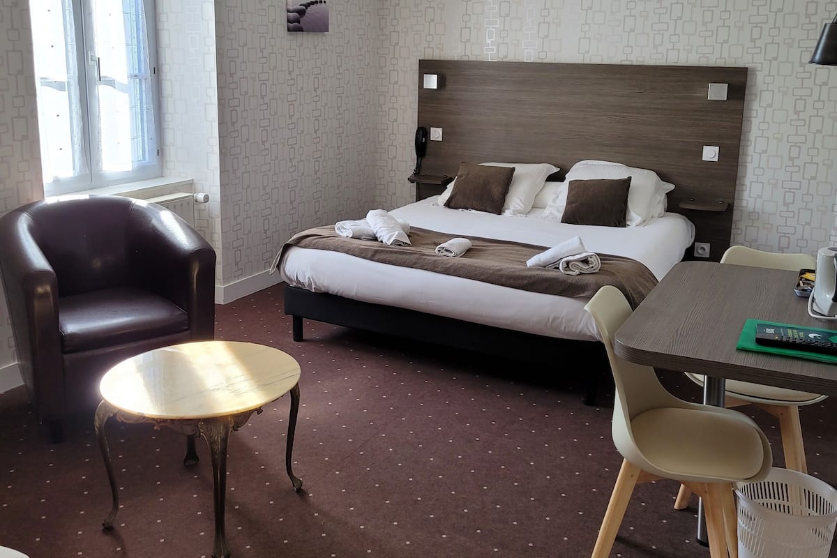 Quarto de hotel com cama, cadeira, mesa pequena e escrivaninha com cadeira.