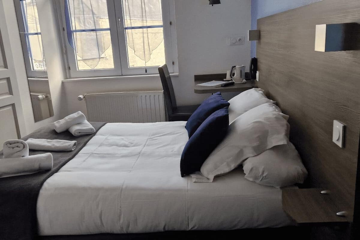 Uma cama bem arrumada com almofadas, uma mesa de cabeceira com abajur e uma janela com cortinas.
