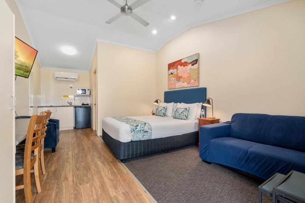 Quarto compacto com cama, sofá azul, mesa de jantar com cadeiras, pequeno espaço de cozinha e ventilador de teto.