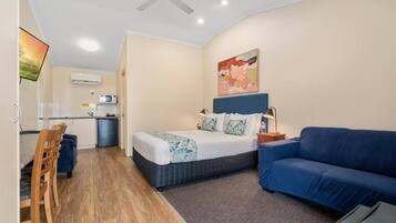 Quarto compacto com cama, sofá azul, mesa de jantar com cadeiras, pequeno espaço de cozinha e ventilador de teto.