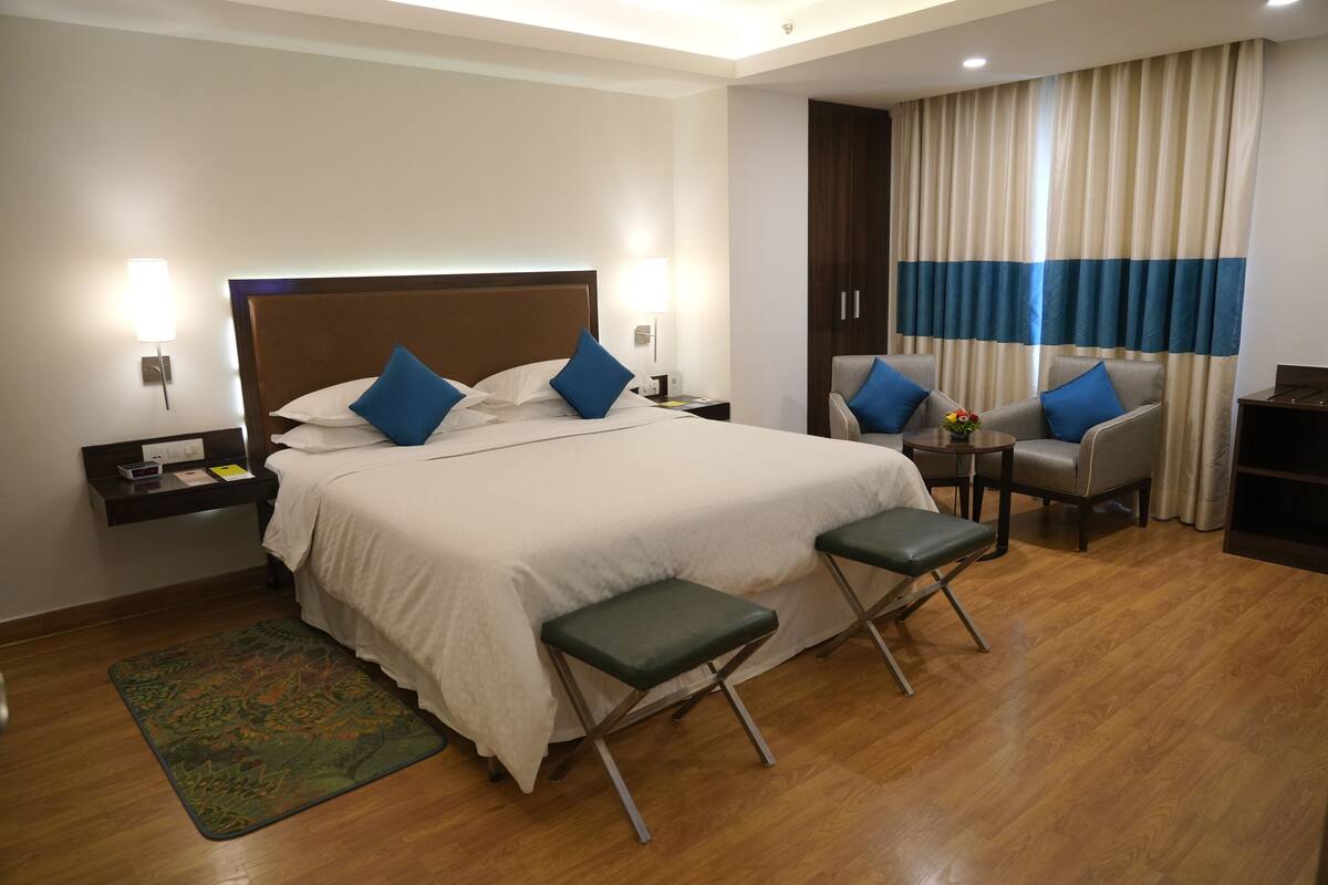 Quarto de hotel com uma cama grande, duas mesas de cabeceira, uma escrivaninha com cadeira, uma pequena mesa com duas cadeiras e uma janela com cortinas.