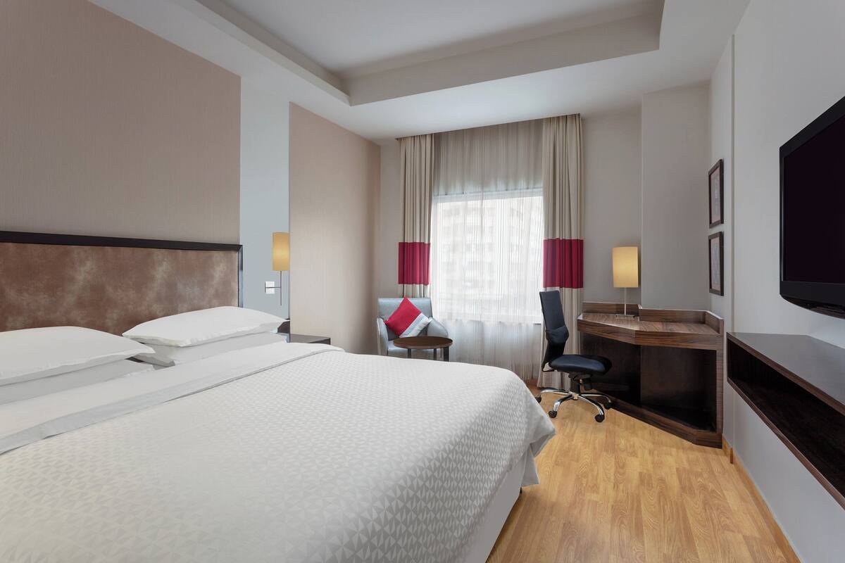 Quarto de hotel com cama grande, escrivaninha, cadeira, televisão e janela com cortinas.