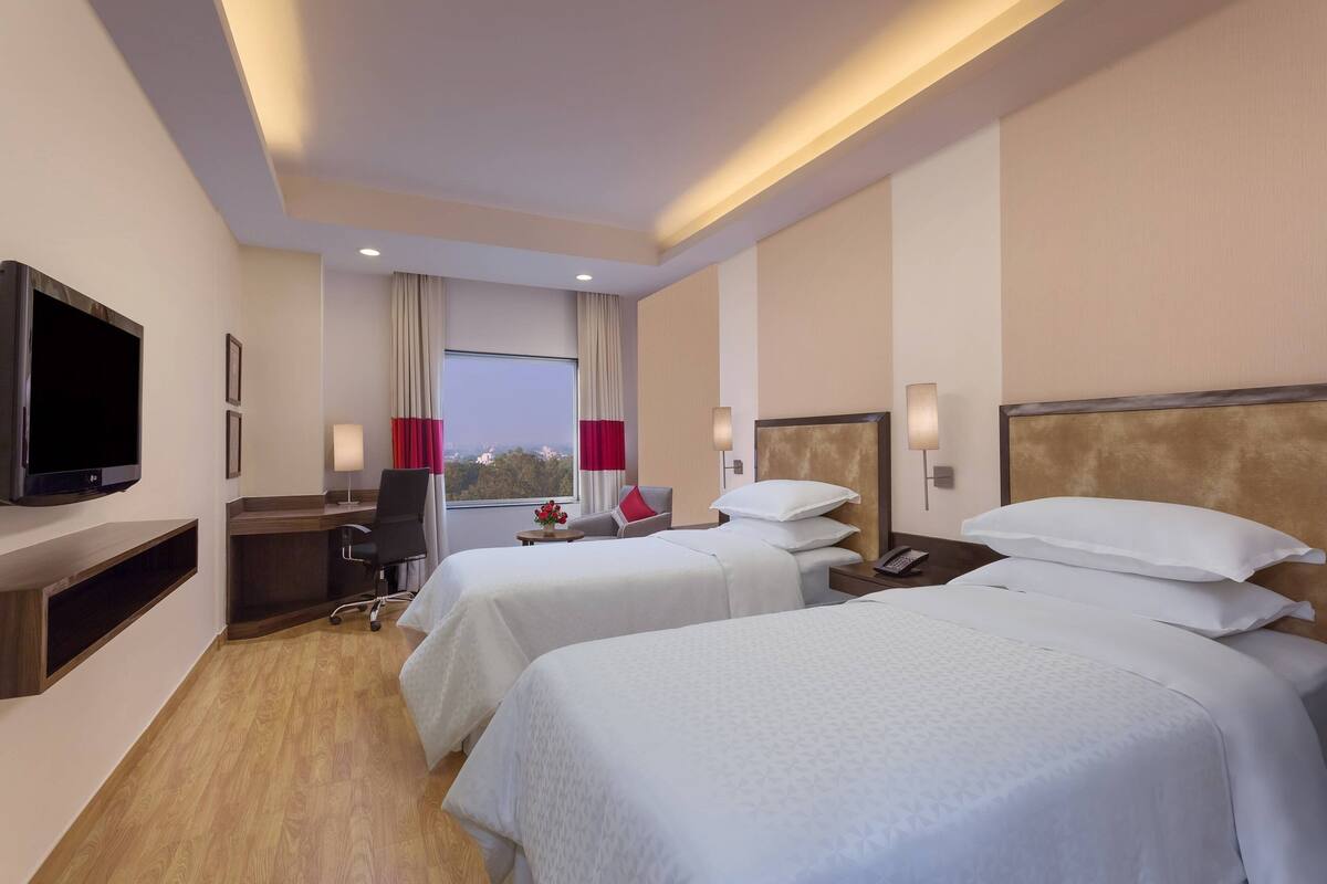 Quarto de hotel com duas camas, uma escrivaninha, televisão e janela com cortinas.