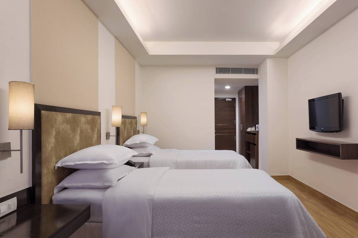 Quarto de hotel com duas camas, televisão, escrivaninha e mesa de cabeceira.