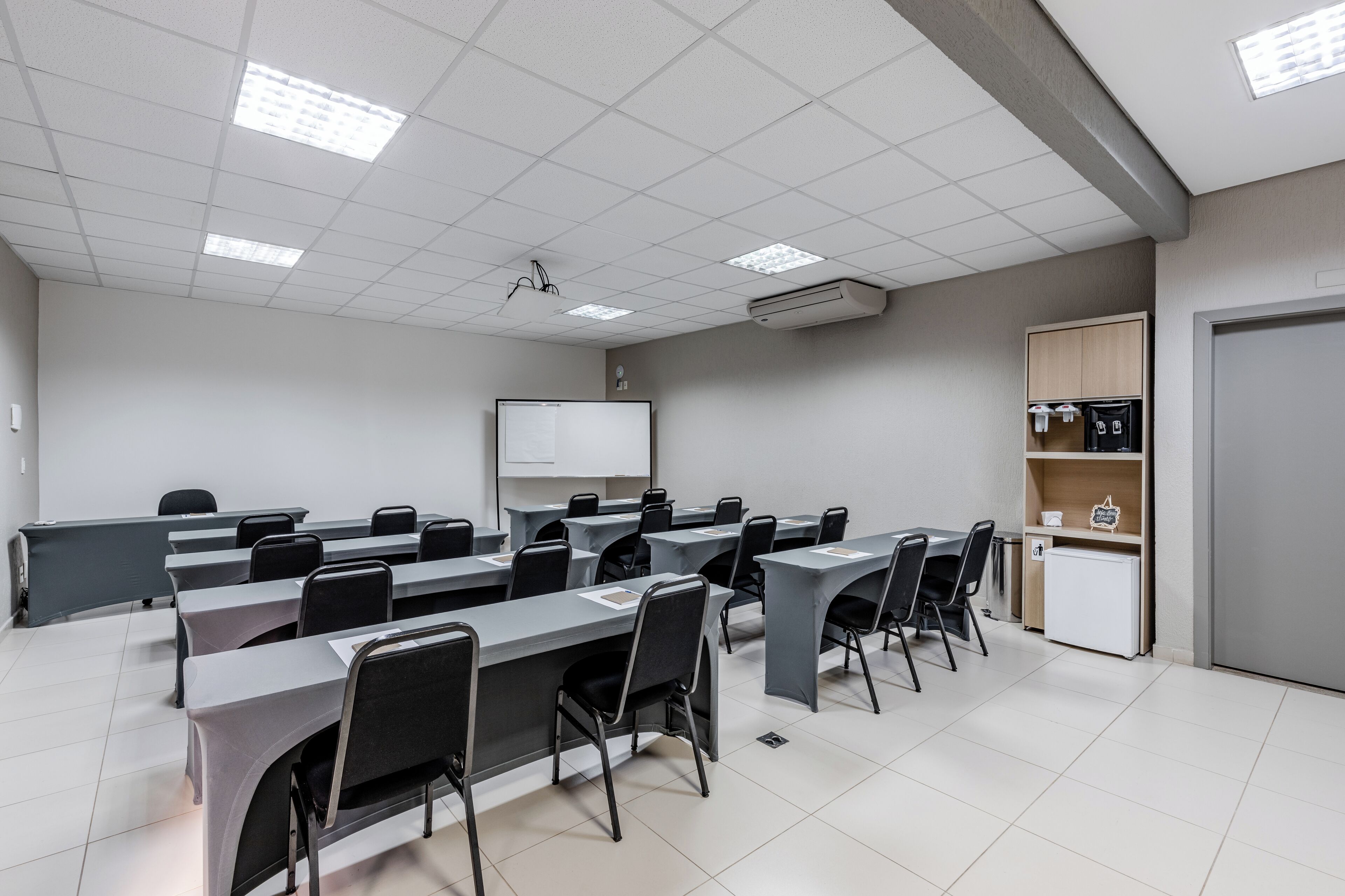Sala de conferências com mesas, cadeiras, quadro branco e pequena cozinha.