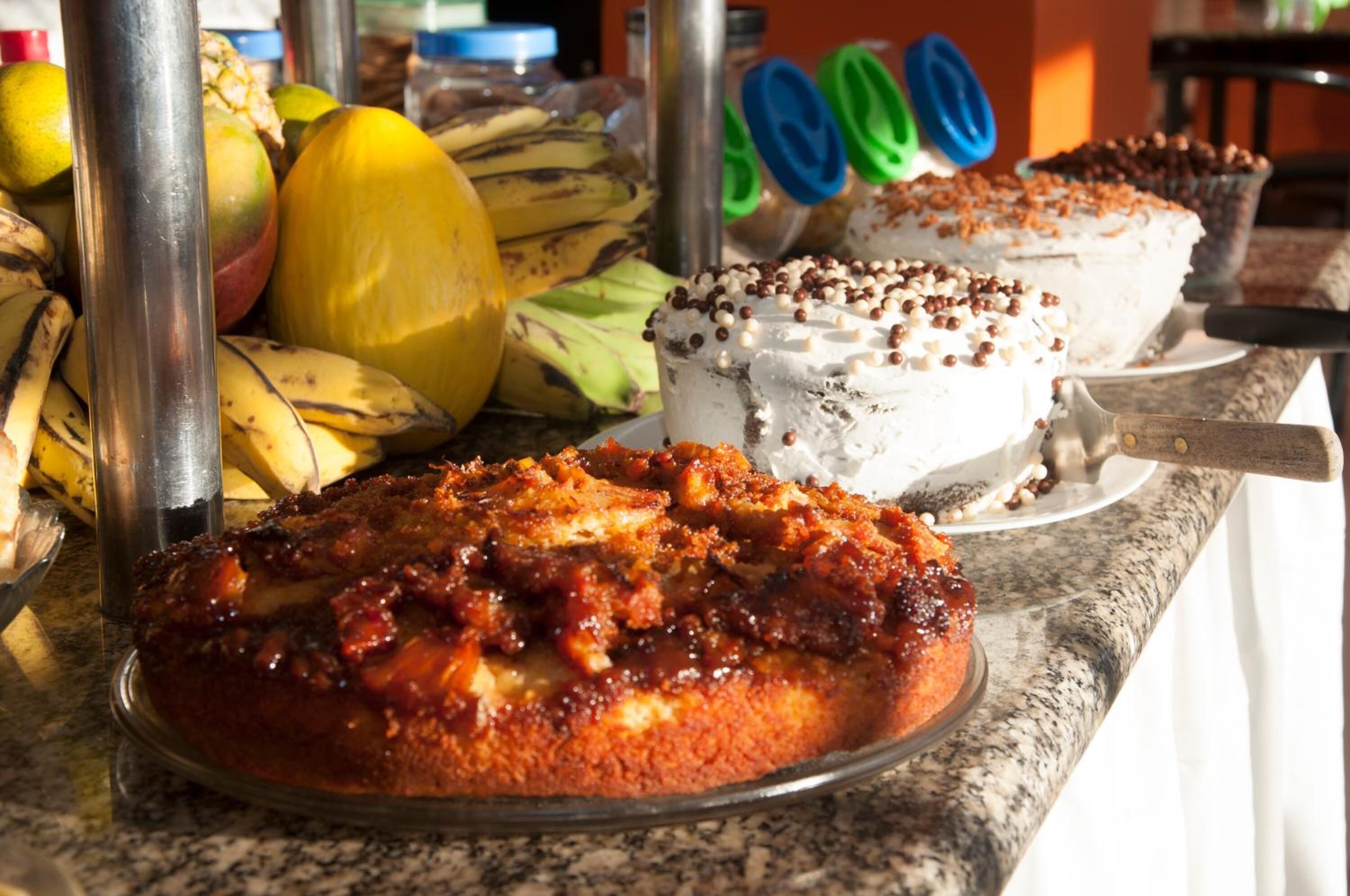 Uma bancada de cozinha com uma variedade de frutas e sobremesas, incluindo um bolo com gotas de chocolate e um bolo coberto de frutas.