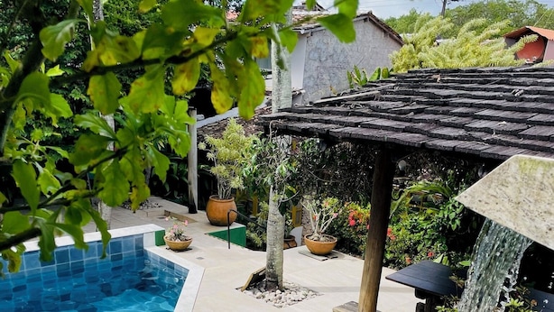 Piscina com cascata, rodeada por plantas em vasos e um pérgula.