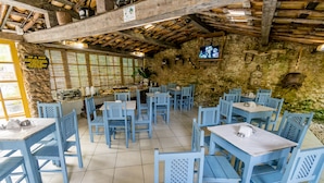 Restaurante rústico com paredes de pedra, vigas de madeira e mobiliário azul. As mesas estão dispostas com toalhas brancas e diversos pratos.