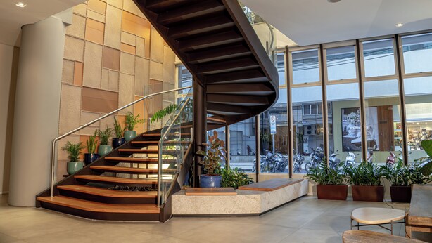 Escadaria em espiral com guarda-corrimão de metal, cercada por plantas em vasos, que leva a um espaço no segundo andar com grandes janelas.