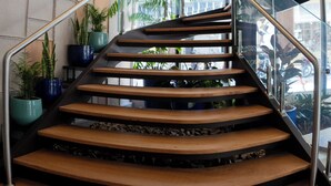 Escadaria em espiral com degraus de madeira e corrimãos de metal, que dá acesso a um segundo andar com grandes janelas e plantas em vasos.