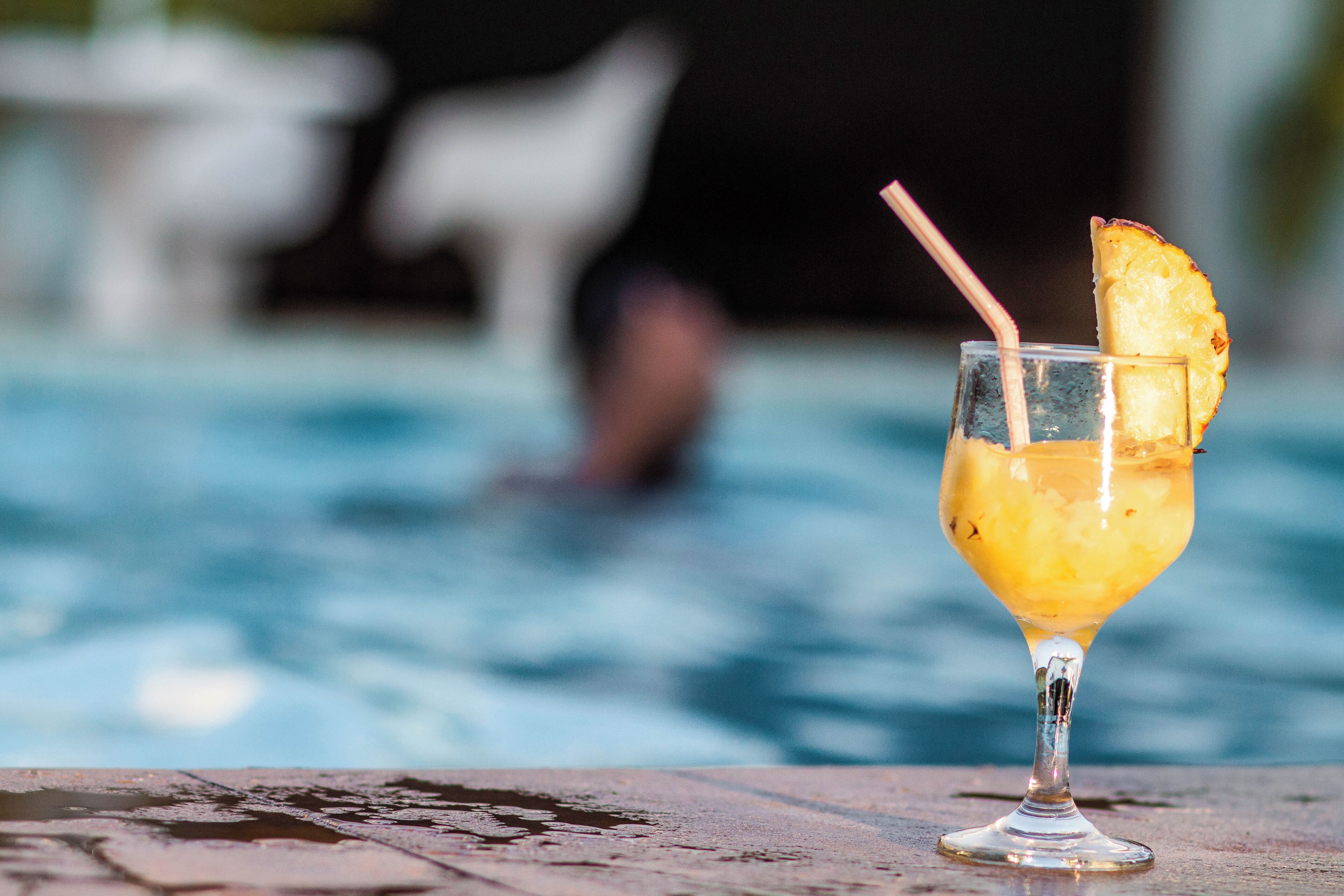Um copo de cocktail com canudo e uma fatia de abacaxi na borda, apoiado sobre uma superfície perto de uma piscina.