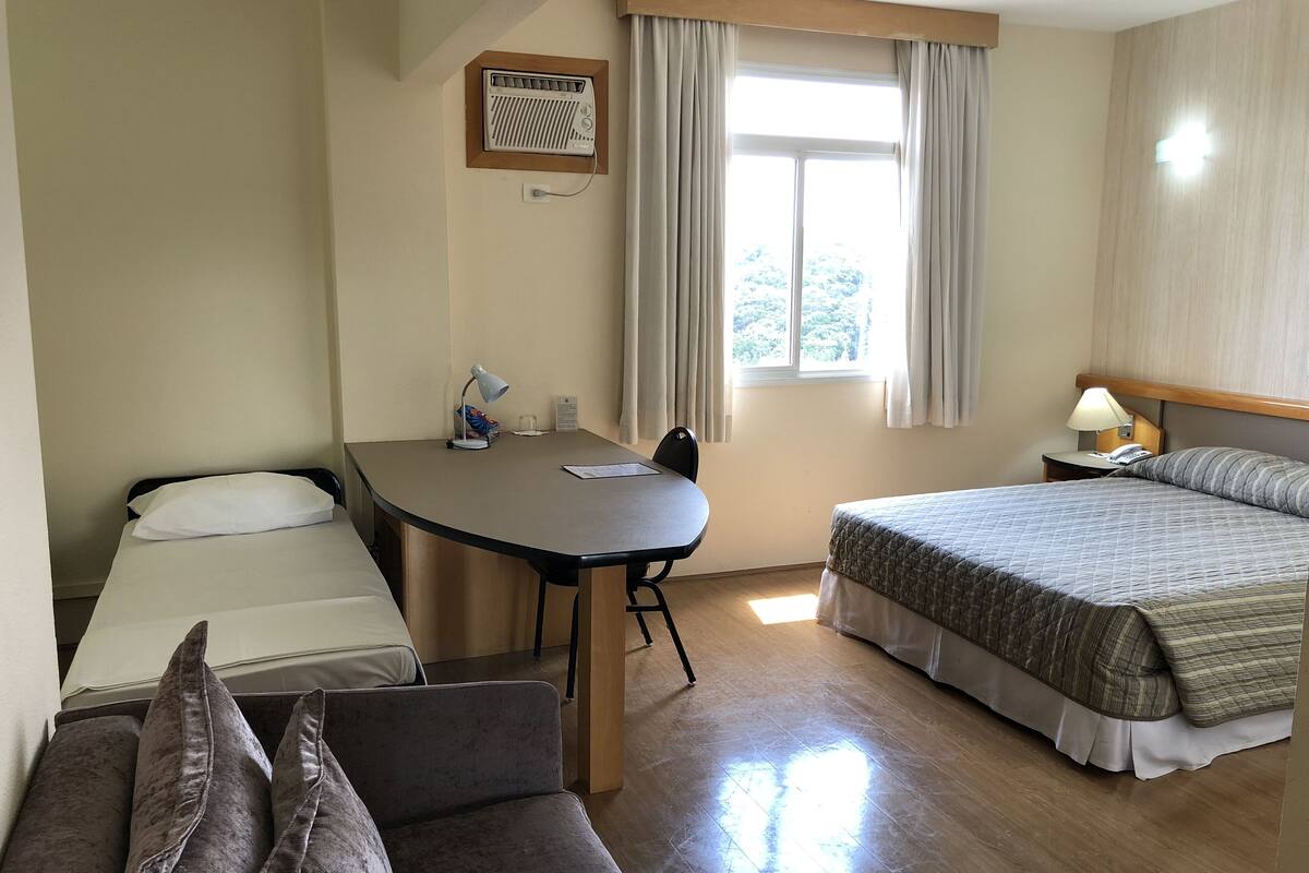 Quarto de hotel com cama, escrivaninha com luminária, cadeira, mesa pequena e janela com cortinas.