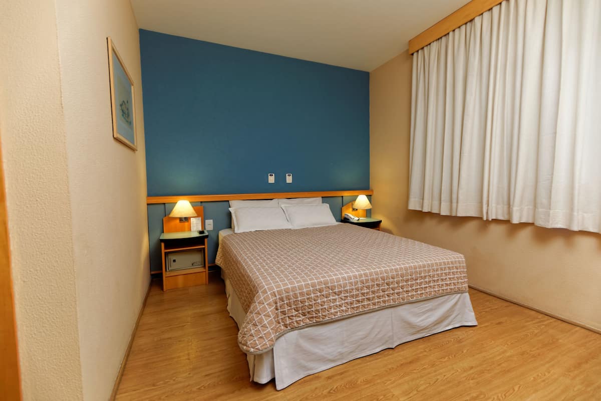 Quarto com cama, duas mesas de cabeceira, um criado-mudo, uma luminária na parede e uma janela com cortinas.