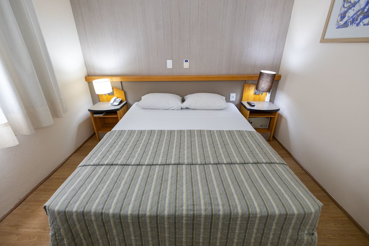 Quarto de hotel com uma cama grande, duas mesas de cabeceira com luminárias, um telefone e um interruptor de luz fixado na parede.
