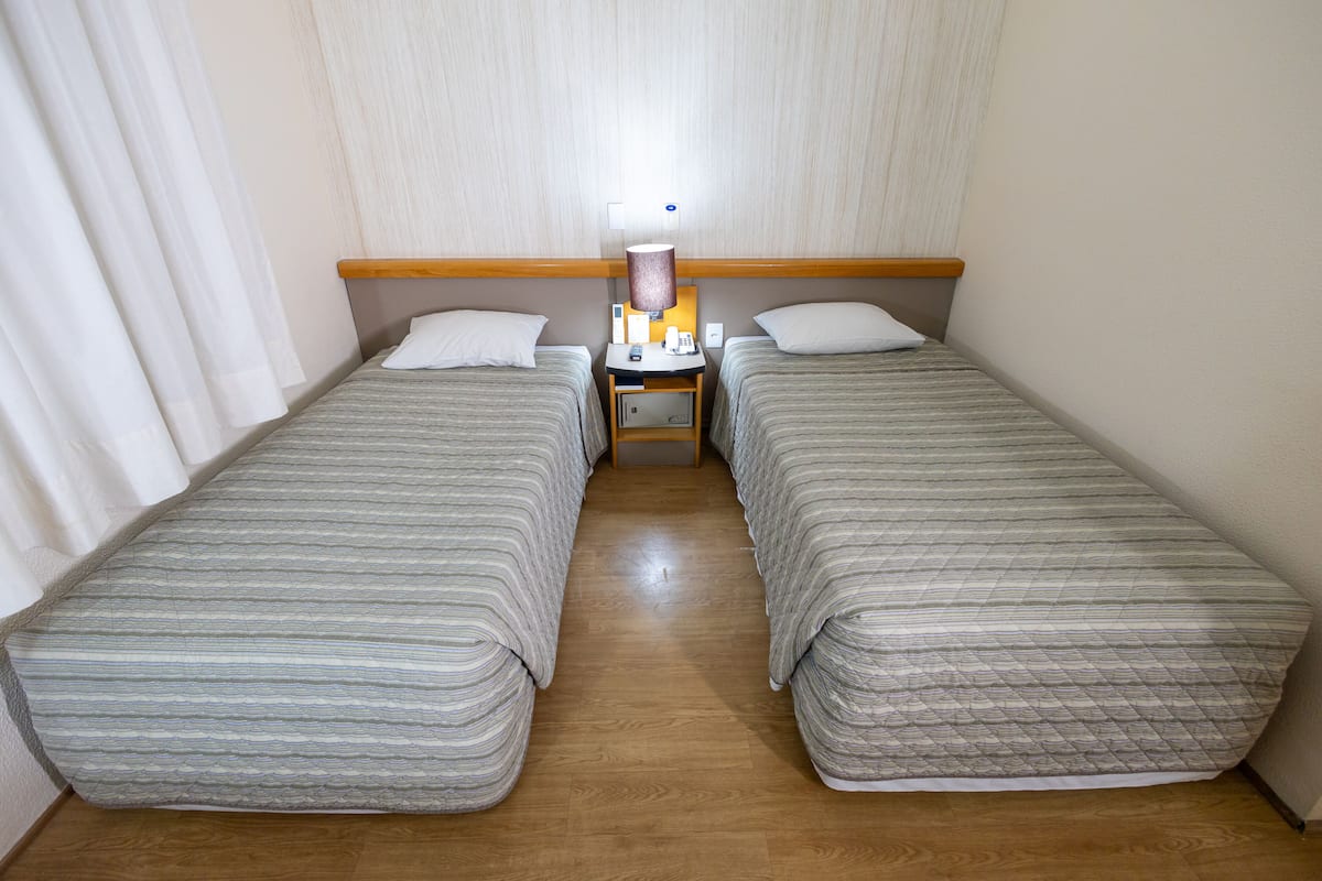 Quarto de hotel com duas camas de solteiro, uma mesinha de cabeceira com abajur e cabeceira de madeira.