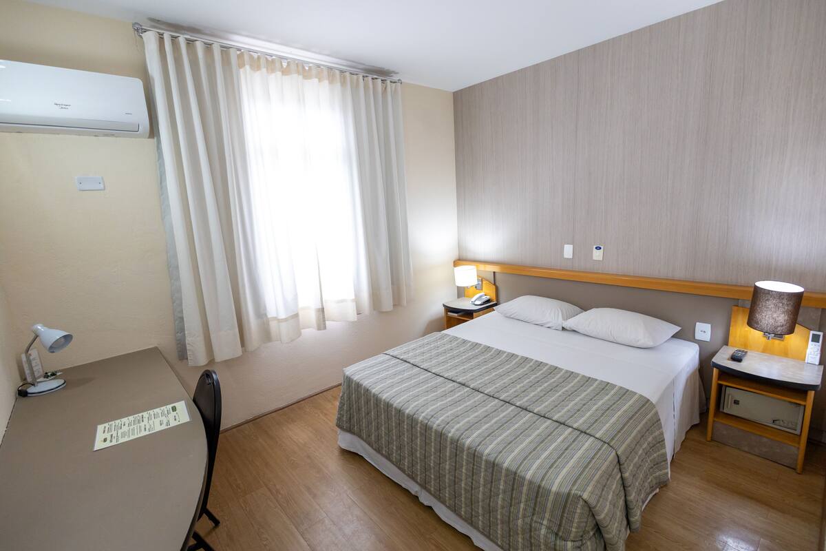Quarto de hotel com cama, mesas de cabeceira, escrivaninha com abajur, cadeira e janela com cortinas.