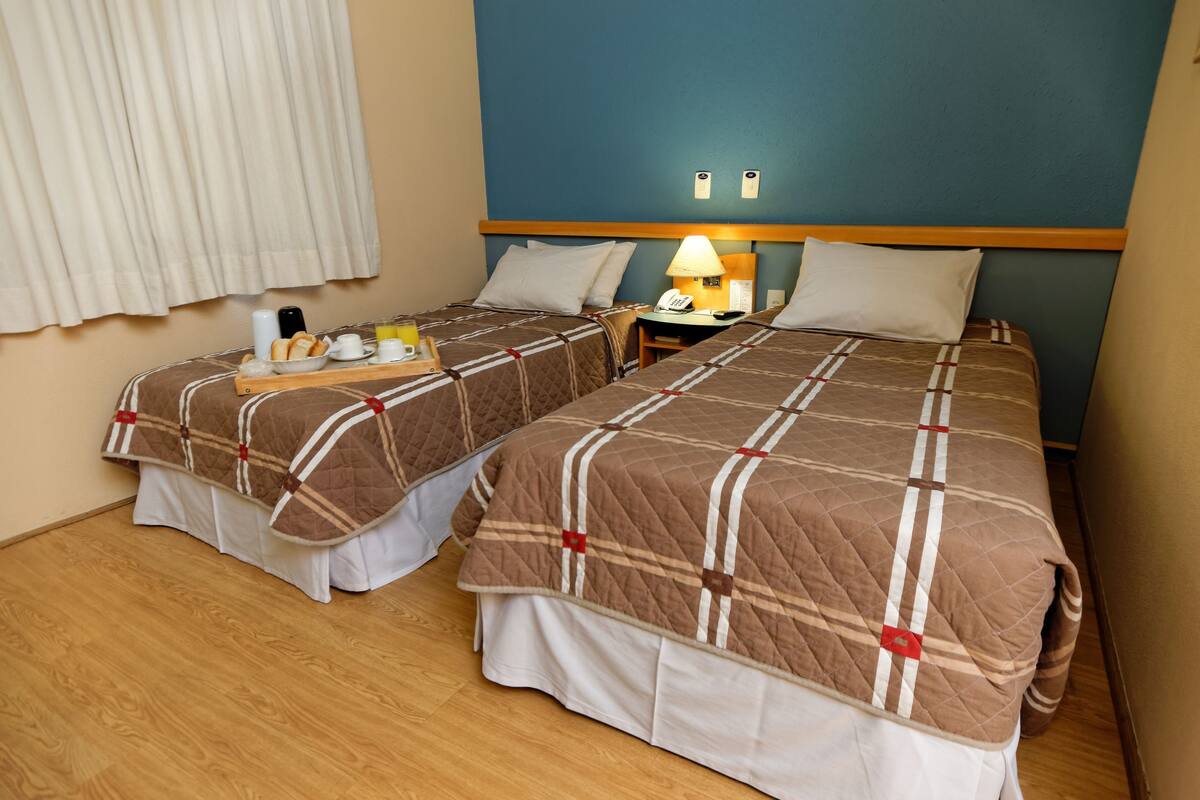 Quarto de hotel com duas camas, uma mesa de cabeceira com abajur, uma pequena mesa com itens de café da manhã e uma janela com cortinas.