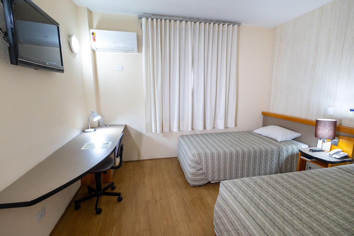 Quarto de hotel com escrivaninha, duas camas, televisão e janela com cortinas.