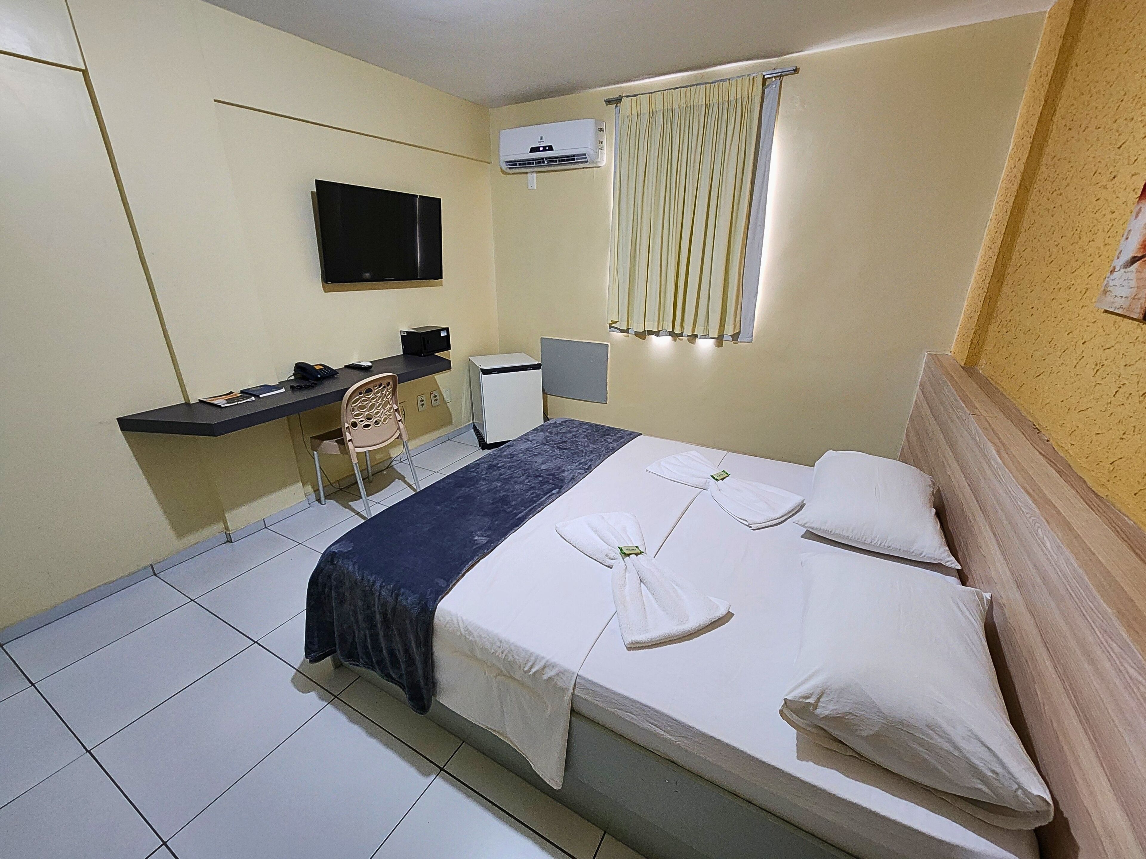 Quarto de hotel com cama, escrivaninha, cadeira, televisão e ar condicionado.