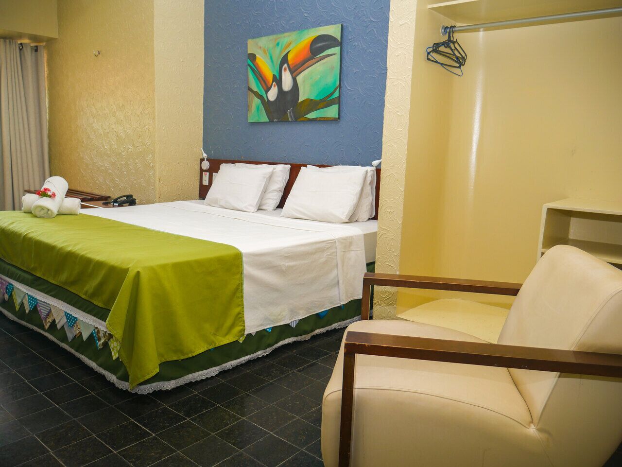Quarto de hotel com cama, uma cadeira, um quadro na parede e porta de banheiro.