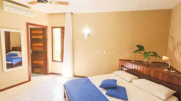Quarto de hotel com cama, ventilador de teto, mesa de cabeceira com abajur e planta em vaso.