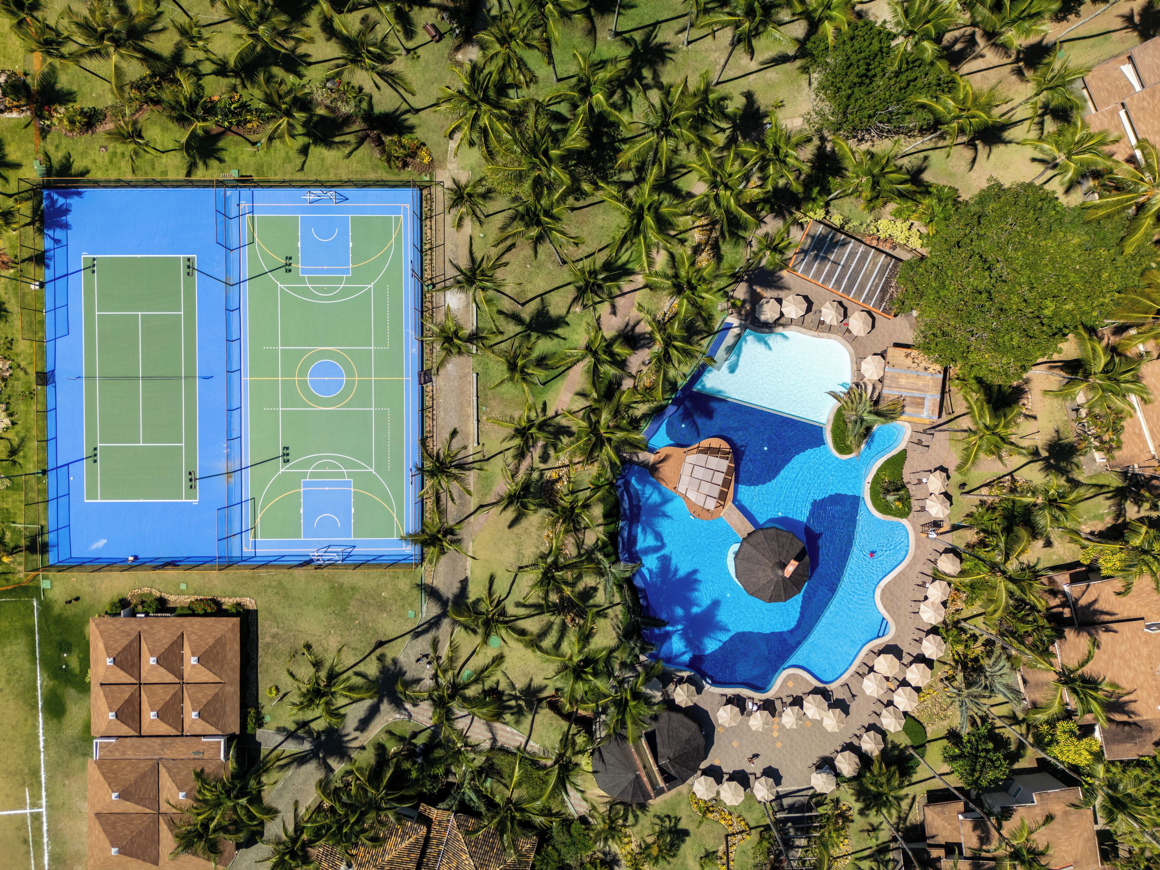 Vista aérea de um resort com quadra de tênis, piscina e palmeiras.